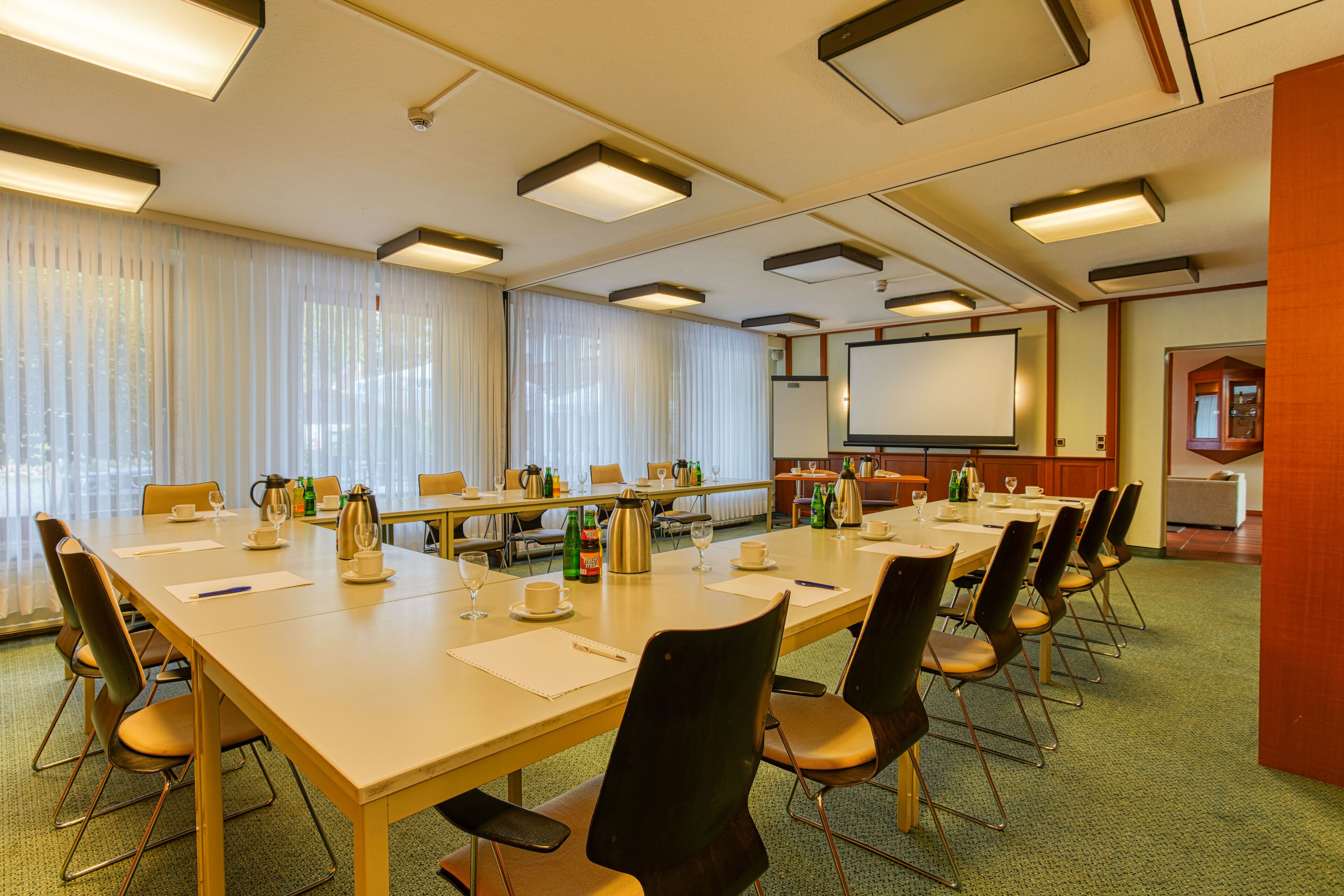 Uma sala de conferência com uma mesa grande, cadeiras, tela de projetor e um espaço para café.