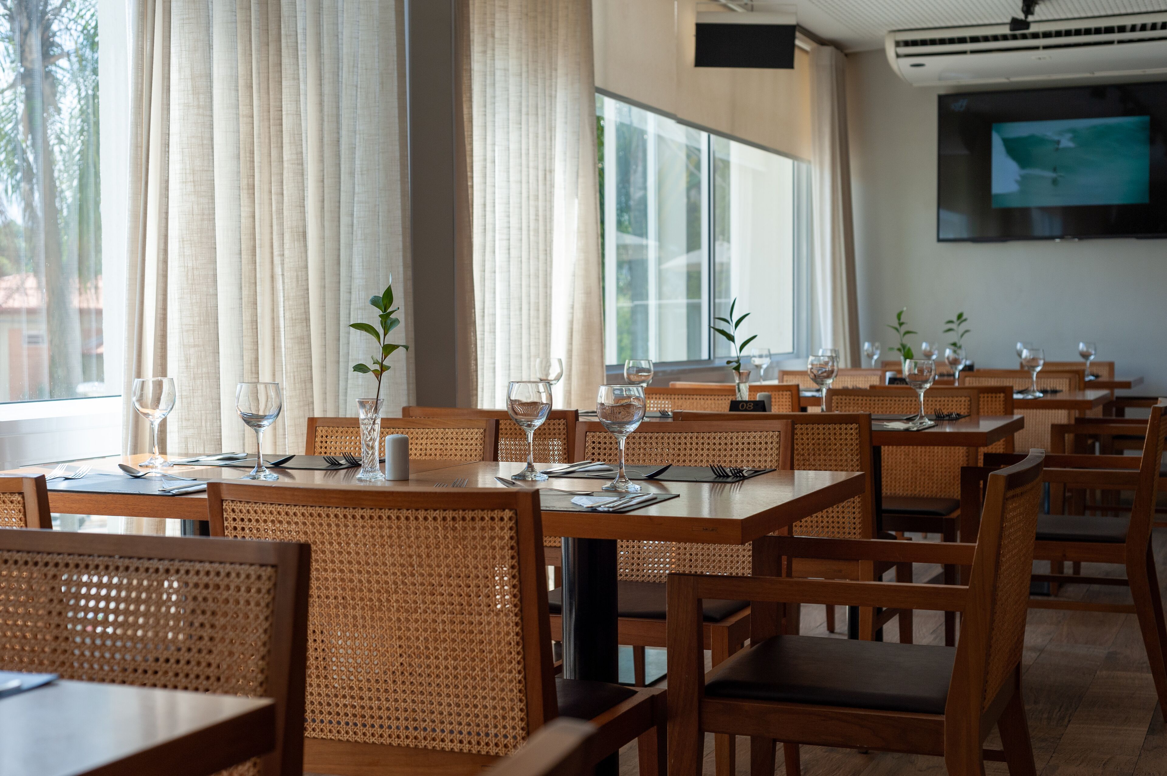 Um restaurante com mesas e cadeiras de madeira, janelas amplas e uma televisão fixada na parede.