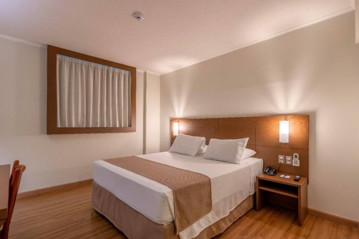 Quarto de hotel com cama, mesa de cabeceira e janela com cortinas.