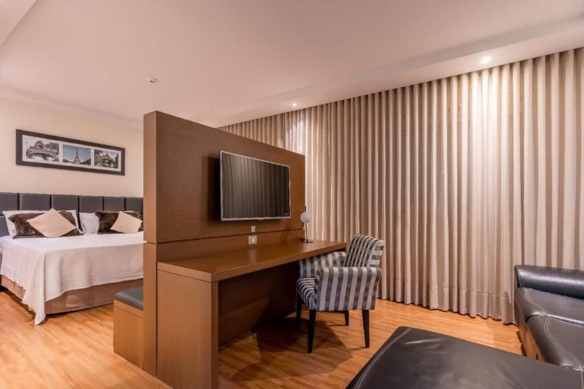 Quarto de hotel com uma cama, uma escrivaninha com televisão, uma cadeira e uma janela grande com cortinas.
