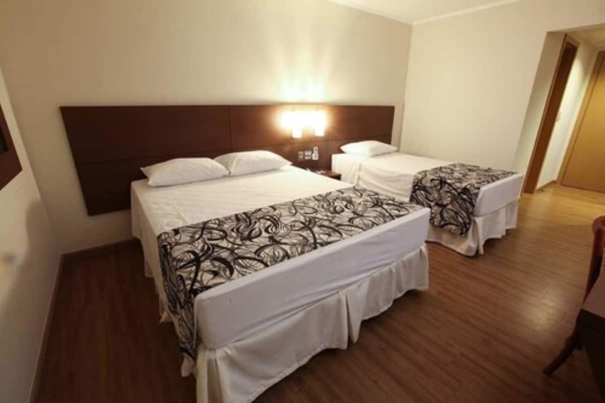 Quarto de hotel com duas camas, cabeceira de madeira e mesa de cabeceira com abajur.