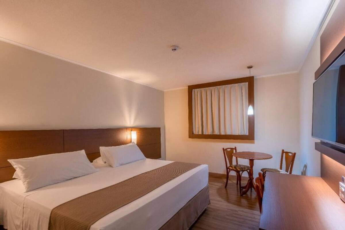 Quarto de hotel com uma cama grande, uma mesa pequena com cadeiras, televisão e uma janela com cortinas.
