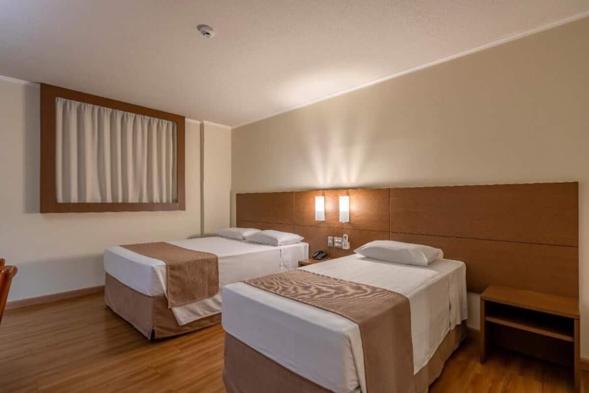 Quarto de hotel com duas camas de solteiro, cabeceira de madeira, mesa de cabeceira, uma cadeira e janela com cortinas.