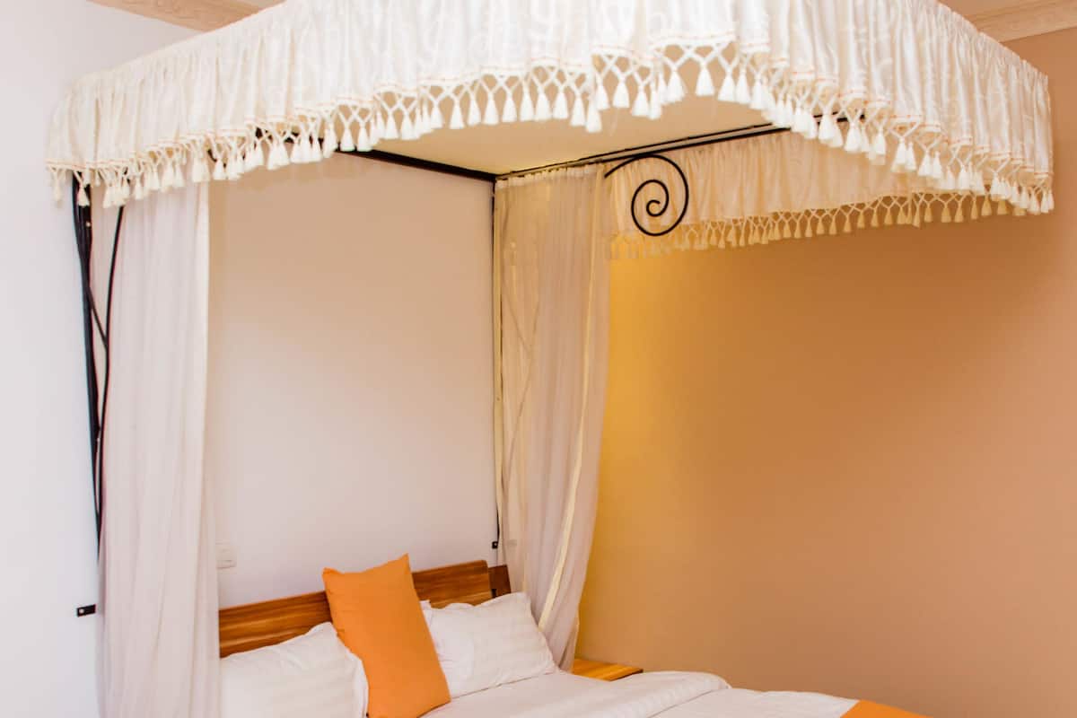 Uma cama com dossel e cortinas, roupa de cama branca e detalhes em laranja, um telefone ao lado da cama e um tapete com padrão floral.