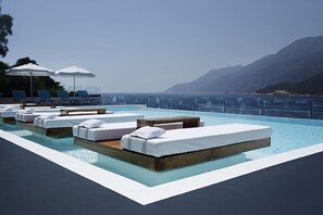 Piscina infinita no terraço com espreguiçadeiras e cadeiras de sol, com vista para o oceano e montanhas.