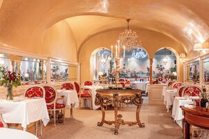 Uma sala de jantar luxuosa com lustres elaborados, cadeiras estofadas em vermelho e toalhas de mesa brancas.