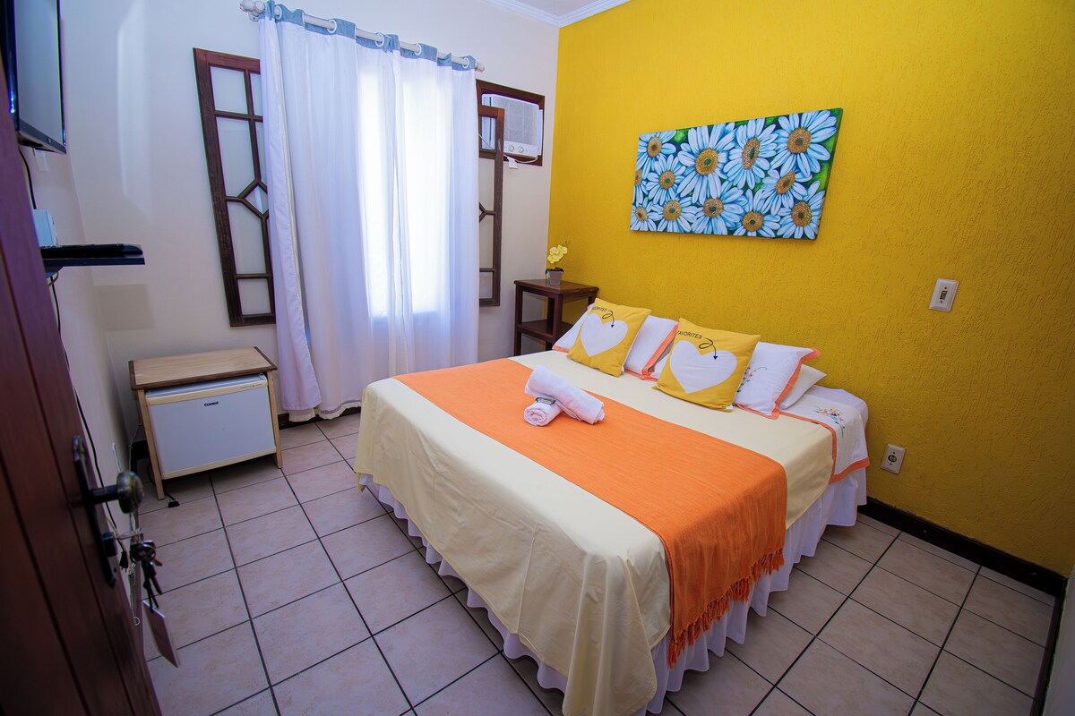 Quarto de hotel com uma cama, mesa de cabeceira, uma pequena mesa, um quadro na parede e uma janela com cortinas.