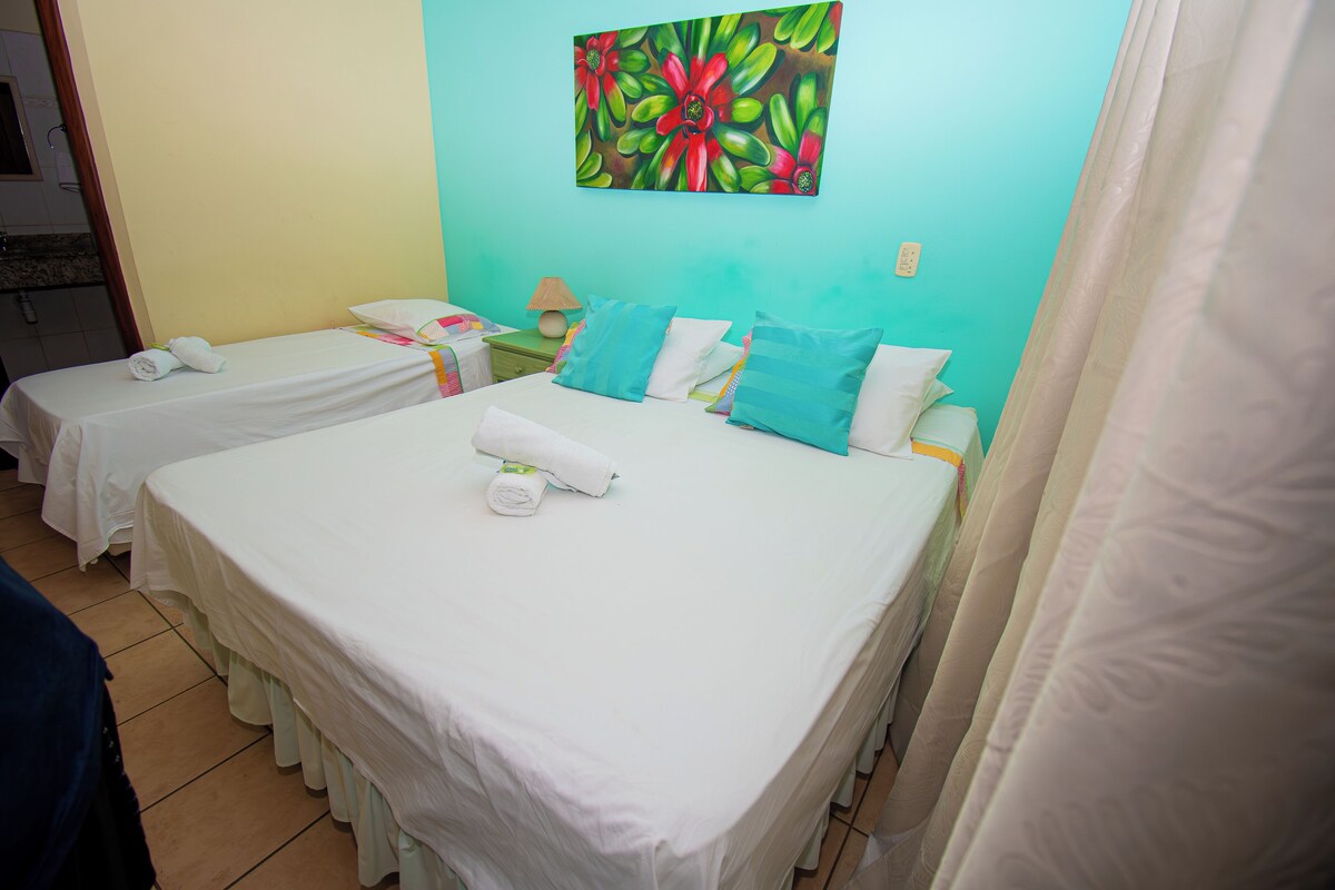 Quarto de hotel com duas camas de solteiro, cada uma com roupa de cama branca e um travesseiro. Há uma pintura de flores na parede acima das camas.