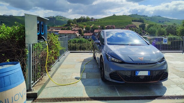 Um carro prateado estacionado em uma estação de carregamento elétrico, com uma vista panorâmica de casas e colinas ao fundo.