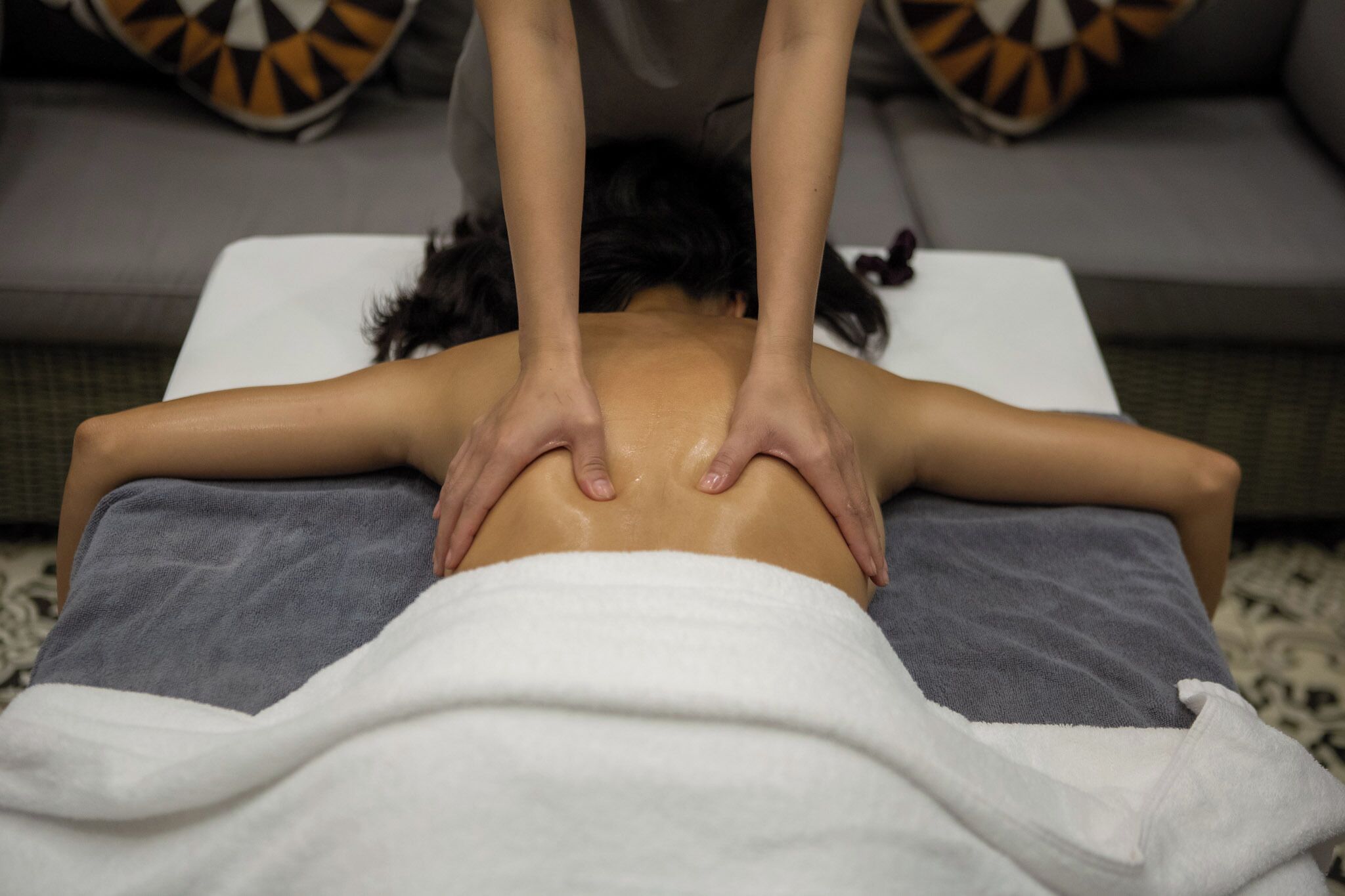 Uma pessoa recebe uma massagem nas costas em uma mesa de massagem, com toalhas e um sofá ao fundo.