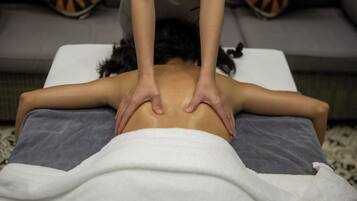 Uma pessoa recebe uma massagem nas costas em uma mesa de massagem, com toalhas e um sofá ao fundo.