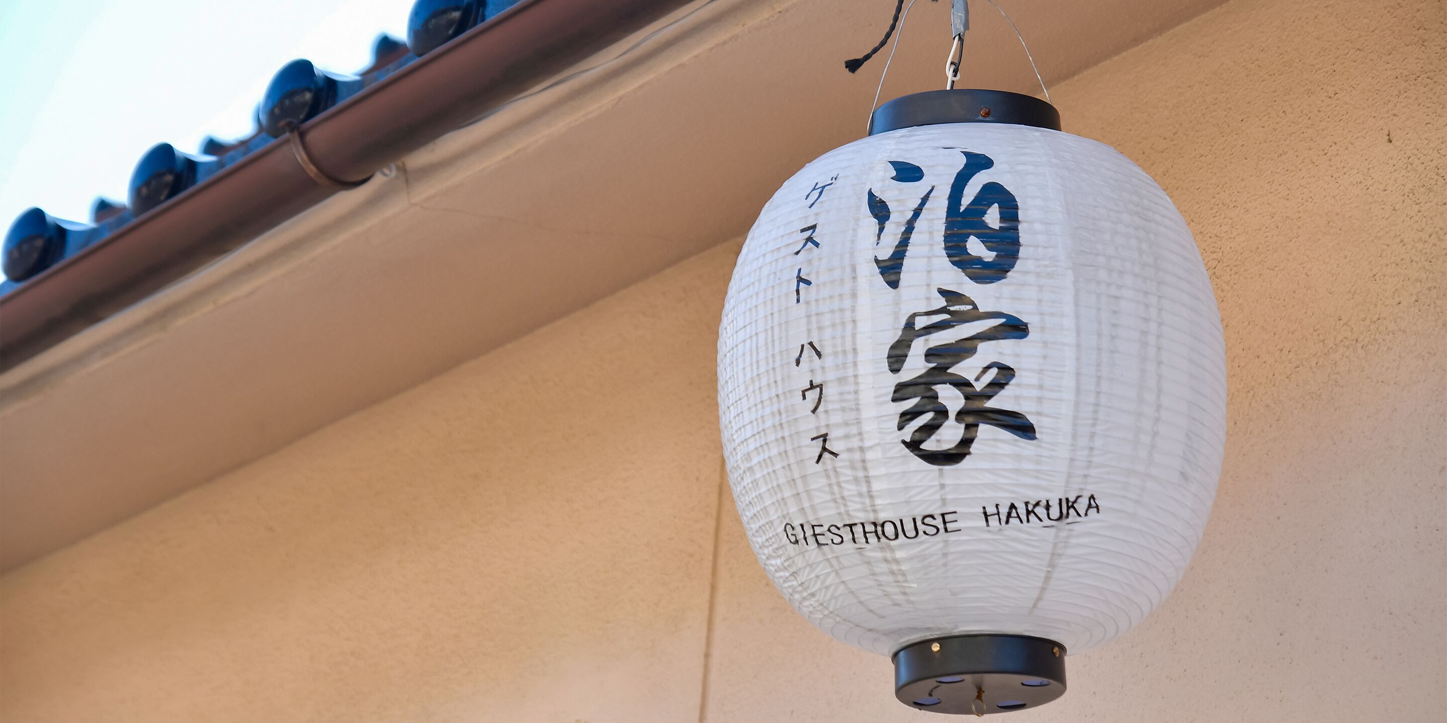 Um candeeiro branco com caracteres japoneses e o texto "GIESTHOUSE HAKUYA" pendurado no teto.