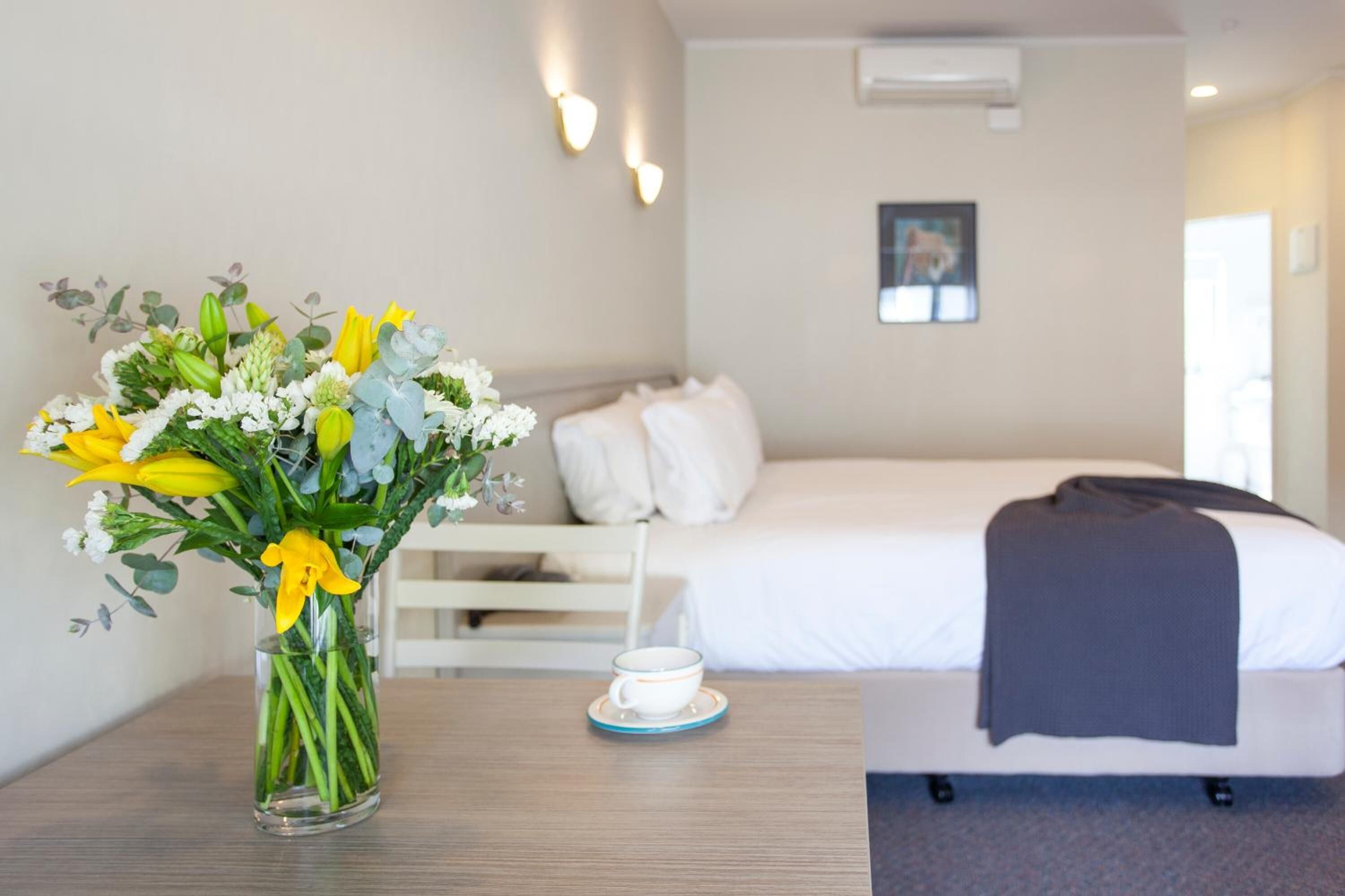 Quarto de hotel com cama, mesa de cabeceira com um vaso de flores, uma chávena e um quadro na parede.