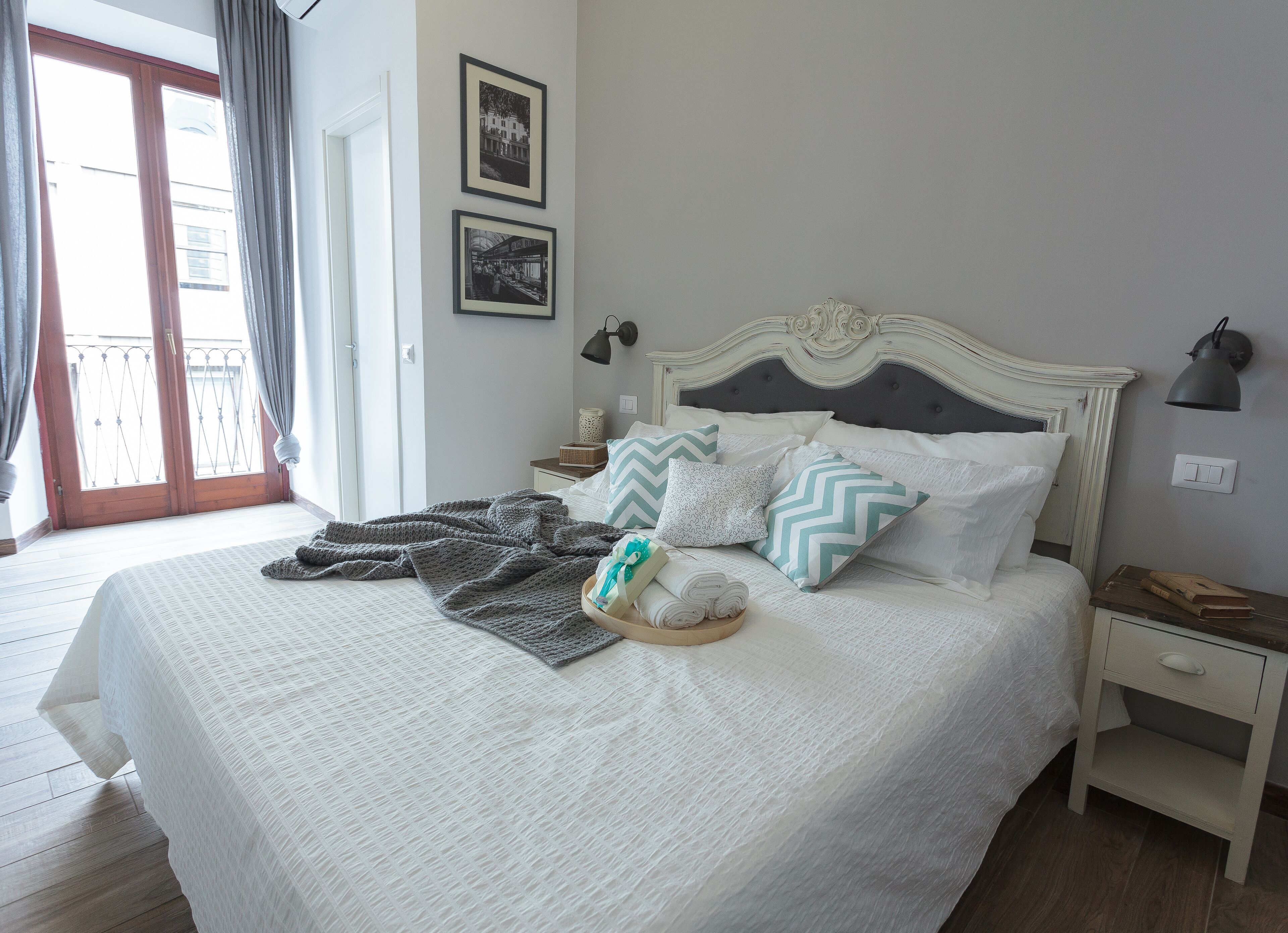 Cama bem arrumada com edredom branco e várias almofadas, mesa de cabeceira com abajur e um quadro na parede.