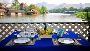 Mesa de jantar posta com pratos, talheres e uma cesta de frutas, com vista para um rio, edifícios tradicionais e montanhas ao fundo.