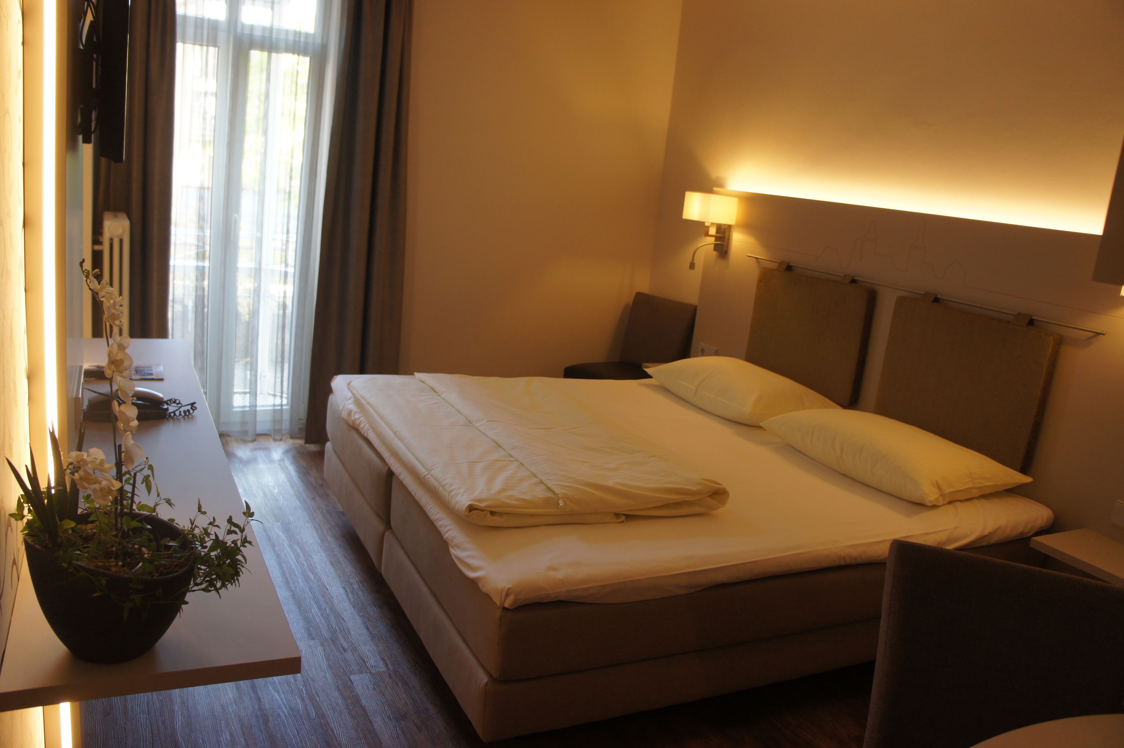 Quarto de hotel com cama, mesa de cabeceira, luminária e planta em vaso.