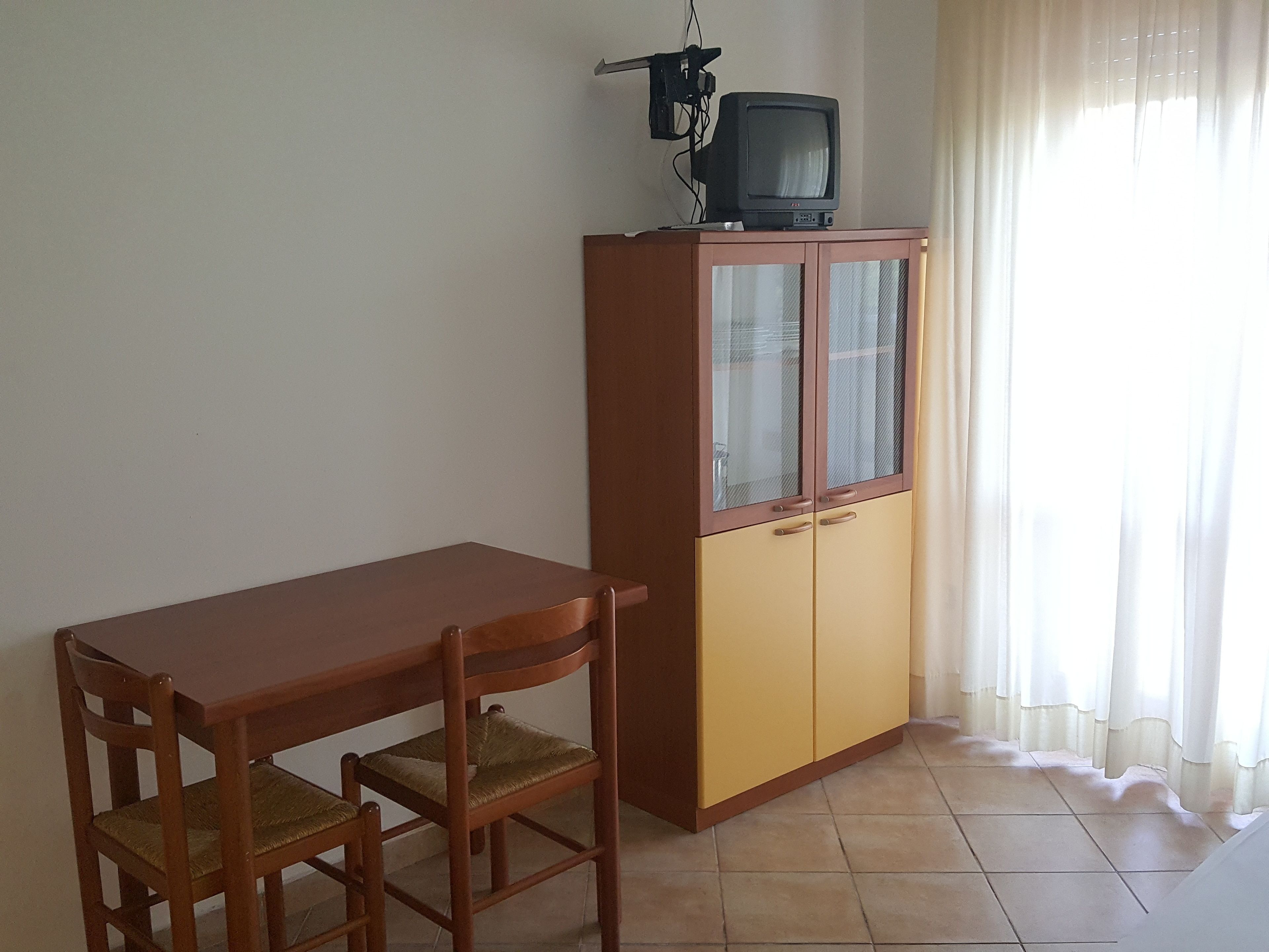 Quarto com mesa e cadeiras de madeira, um armário com portas de vidro e uma televisão em cima.