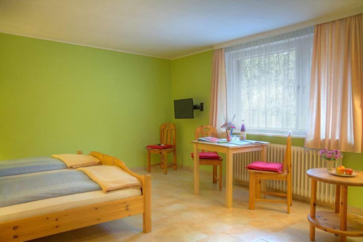 Quarto com cama, escrivaninha, duas cadeiras, radiador e janela com cortinas.