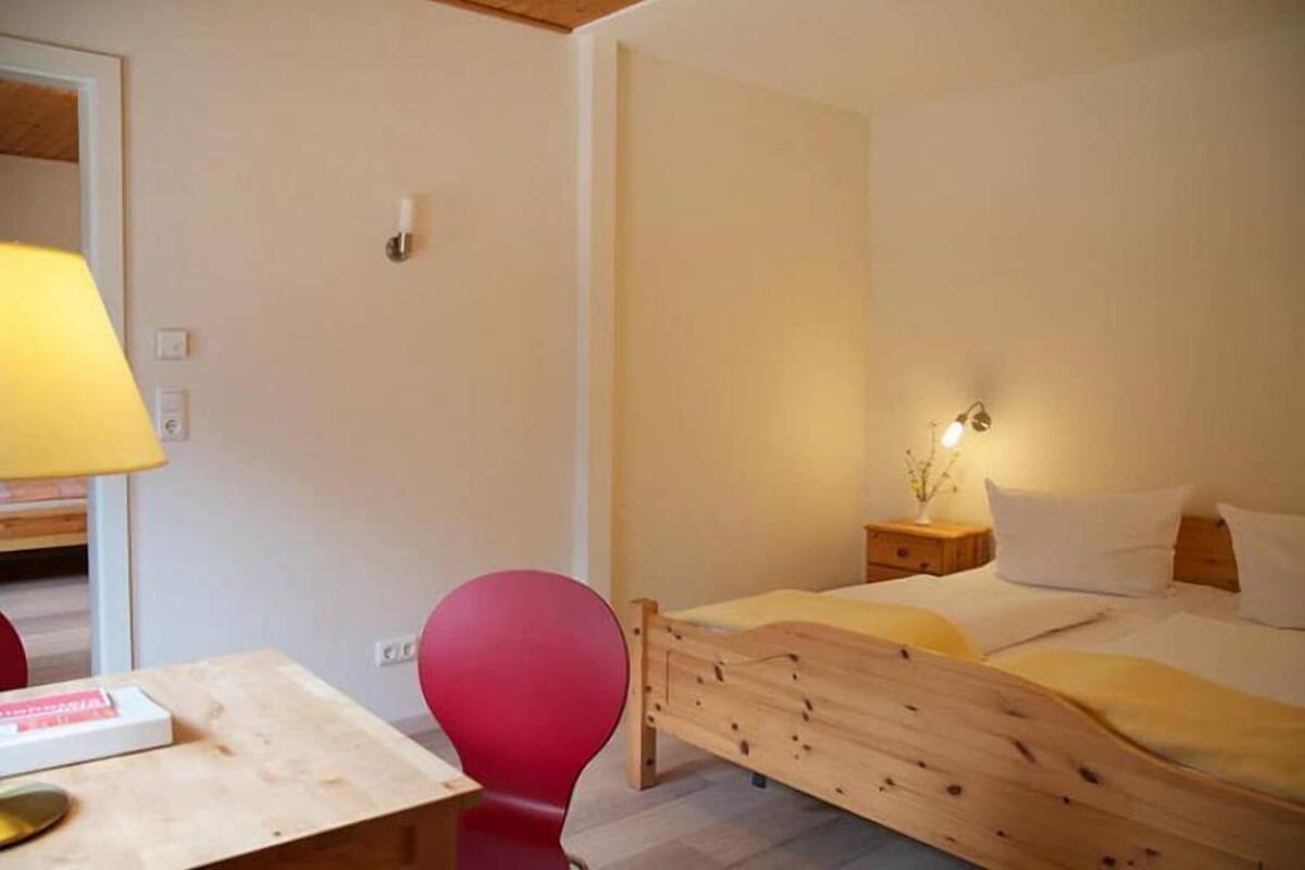 Quarto com cama de madeira, mesa de cabeceira com abajur, uma cadeira vermelha e pequena mesa de cabeceira com uma planta.