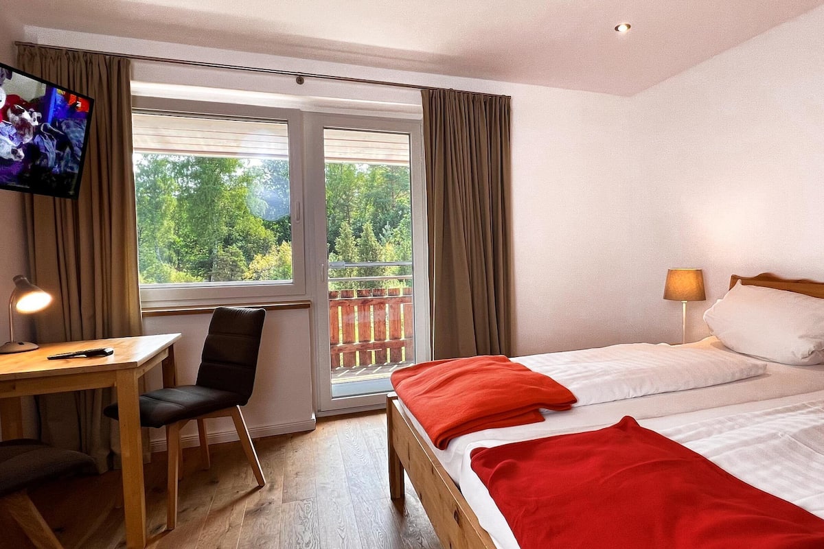 Quarto de hotel com cama, escrivaninha, cadeira, televisão e varanda com cortinas.