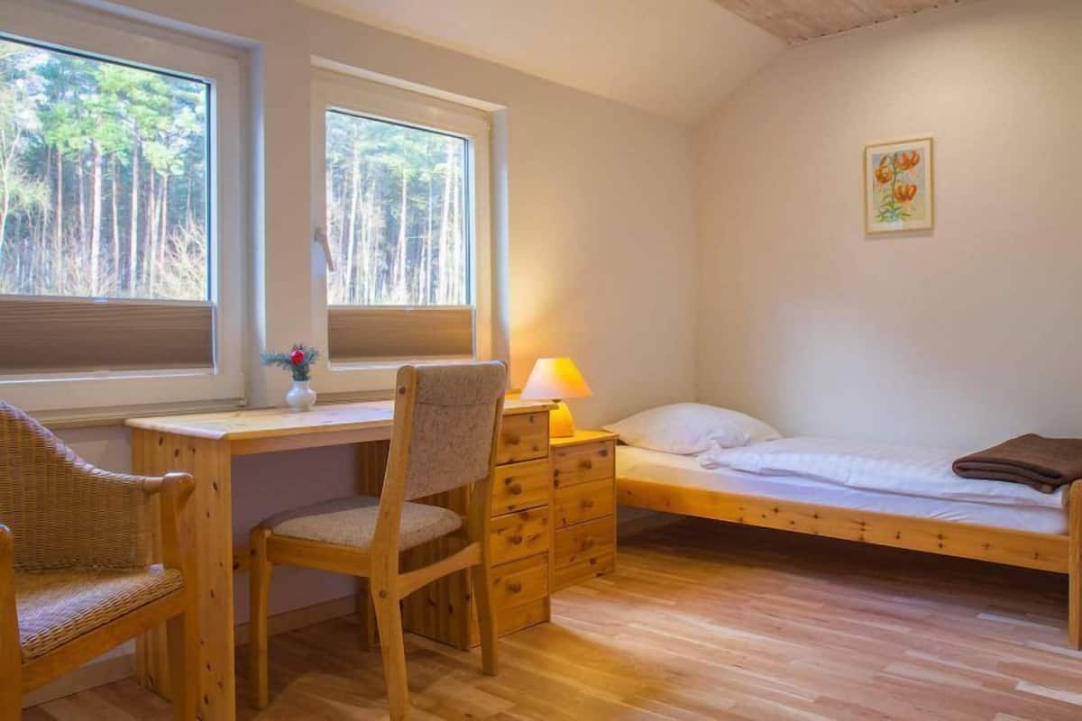 Quarto com escrivaninha de madeira, cadeira e cama. Duas janelas oferecem vista para árvores.