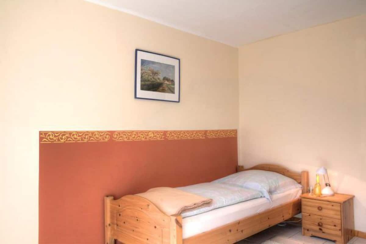 Quarto individual com uma cama e cabeceira de madeira, mesa de cabeceira com abajur e um quadro na parede.