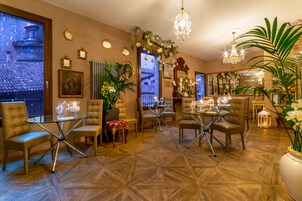 Uma área de refeições com piso em parquet de madeira, mesas redondas e cadeiras. Há plantas decorativas e um candelabro.