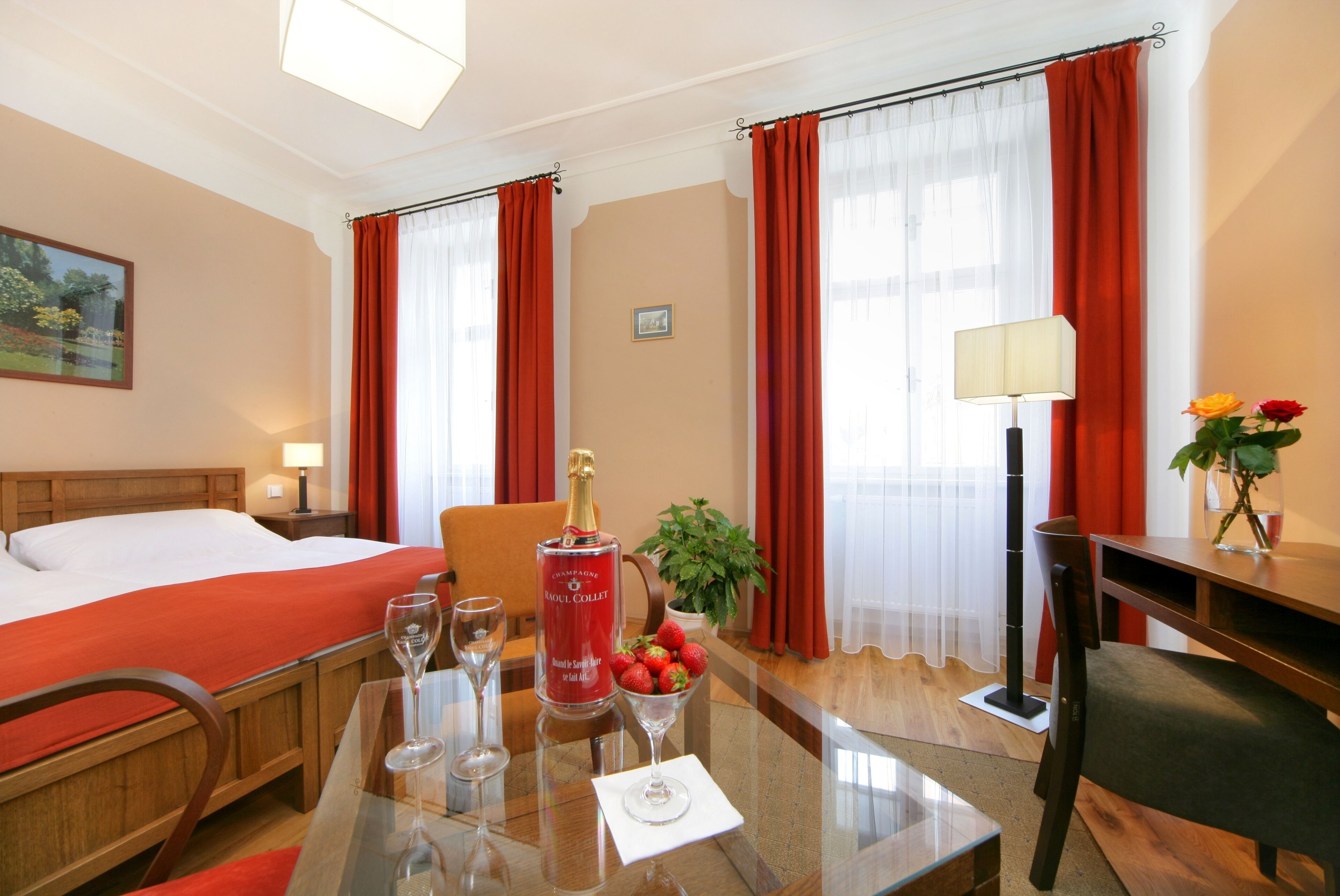 Quarto de hotel com uma cama, uma mesa com champanhe, copos, morangos e uma planta em vaso.