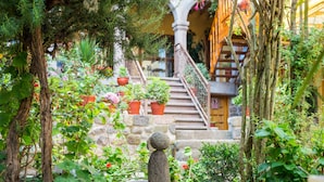 Um jardim com uma escultura de pedra, uma escadaria que leva a uma porta de madeira e diversos vasos de plantas.