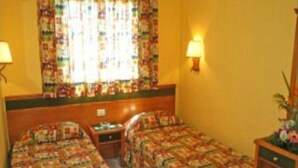 Quarto de hotel com duas camas de solteiro, uma pequena mesa, mesa de cabeceira, abajur e janela com cortinas.