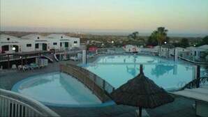 Área de piscina com guarda-sol de palha, espreguiçadeiras e vista para o horizonte urbano.