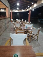 Uma área de refeições ao ar livre com mesas de madeira e cadeiras brancas, sob um telhado coberto com vigas e luminárias aparentes.