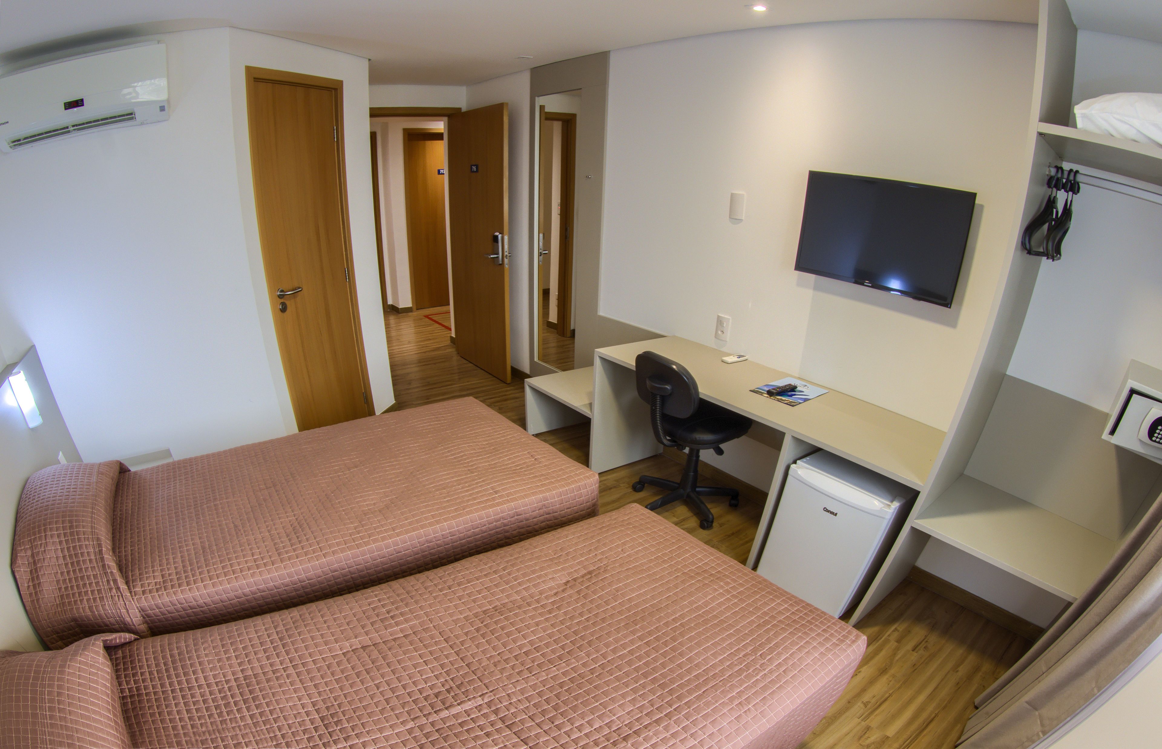 Quarto de hotel com duas camas, uma escrivaninha com cadeira, televisão e um pequeno frigorífico.