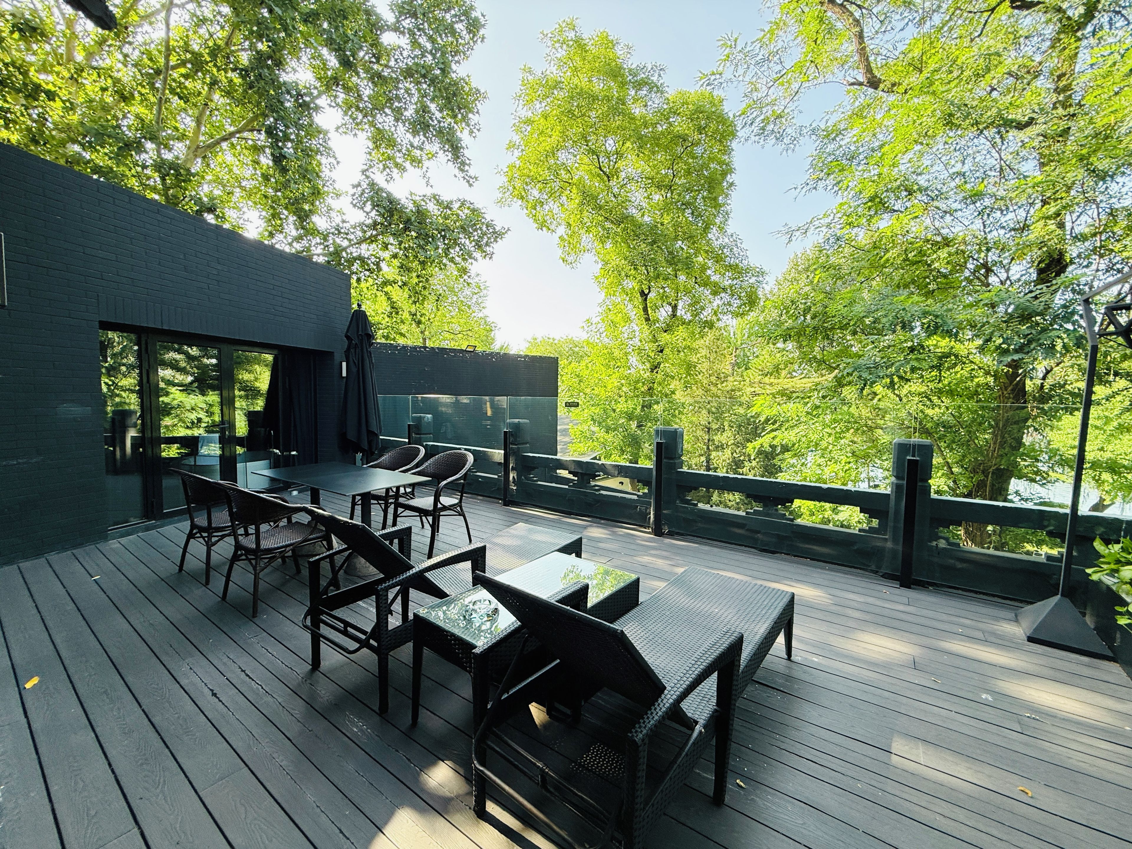 Um deck externo com mesa e cadeiras, cercado por corrimão de madeira e com vista para uma área arborizada.