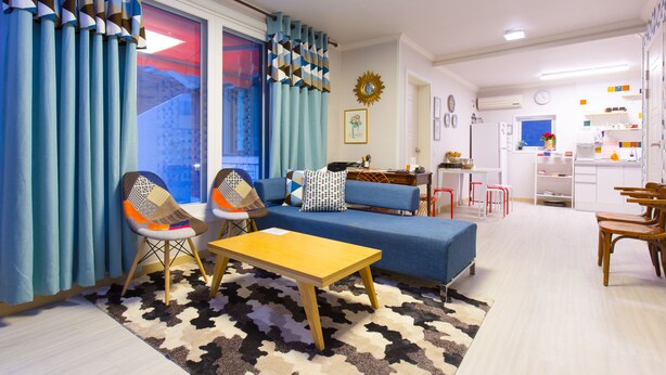 Uma sala de estar moderna com um sofá azul, uma mesa de centro de madeira e cadeiras estampadas. Uma área de cozinha é visível ao fundo, com armários brancos e uma mesa de jantar.