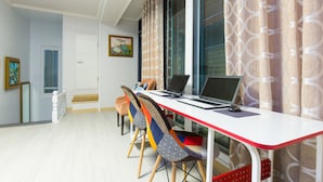 Quarto moderno com escrivaninha branca, duas cadeiras e um laptop. Uma janela grande com cortinas estampadas.