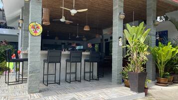 Uma área de bar ao ar livre com estrutura de coluna de pedra, vários arranjos de assentos e ventiladores de teto.