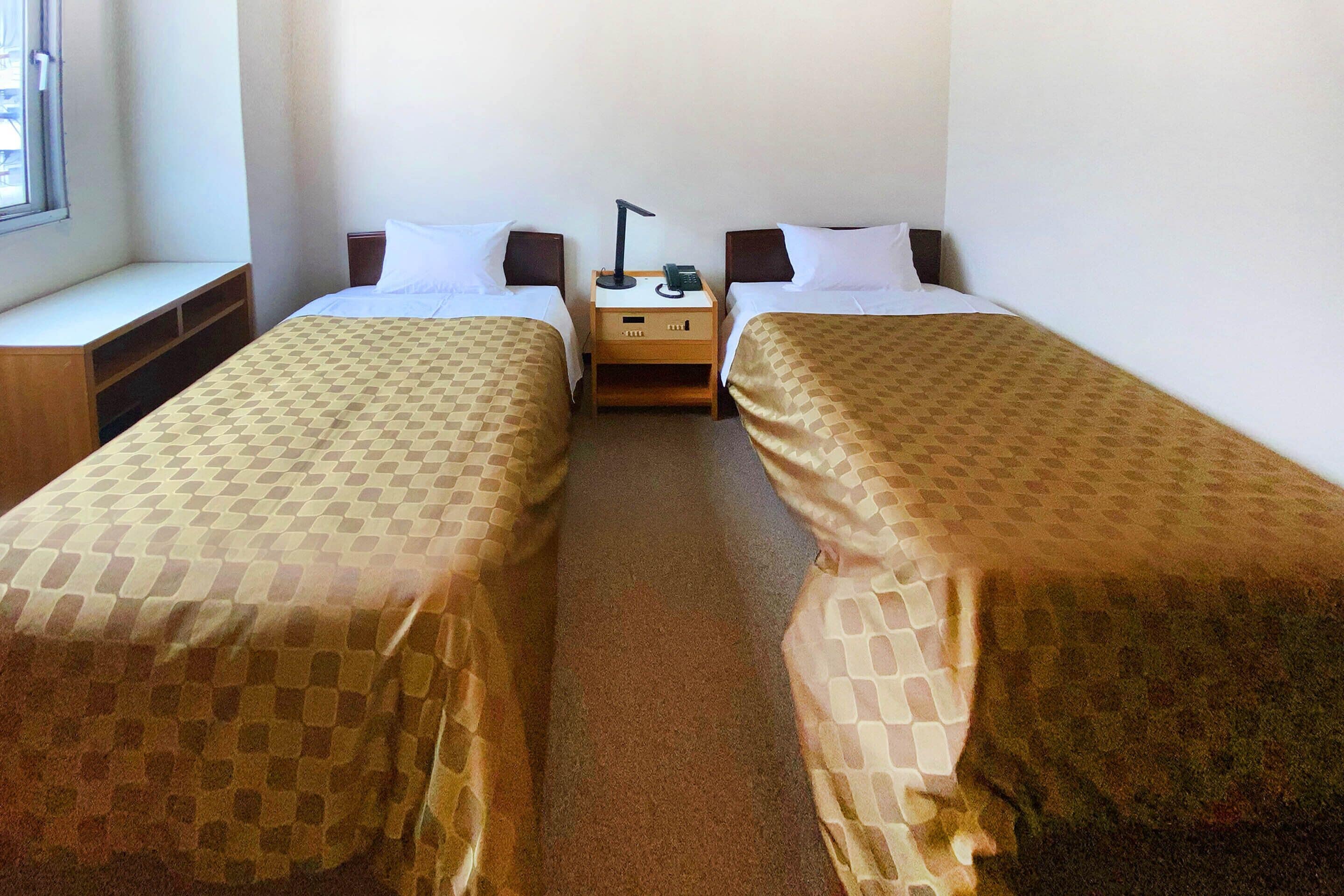 Quarto de hotel com duas camas de solteiro, uma mesa de cabeceira com luminária e uma janela.