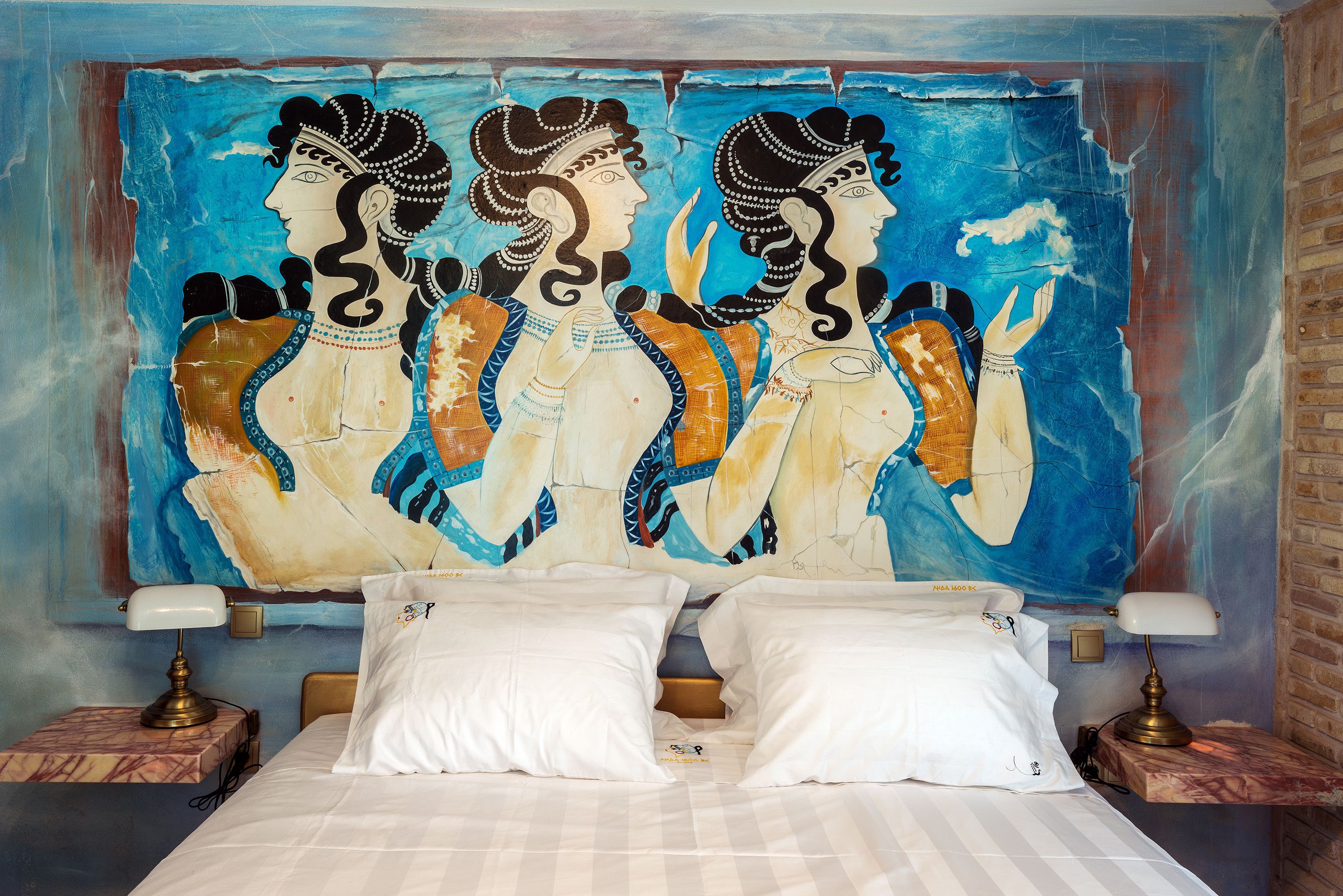 Uma cama com roupa de cama branca, duas mesas de cabeceira com luminárias e um grande mural na parede.