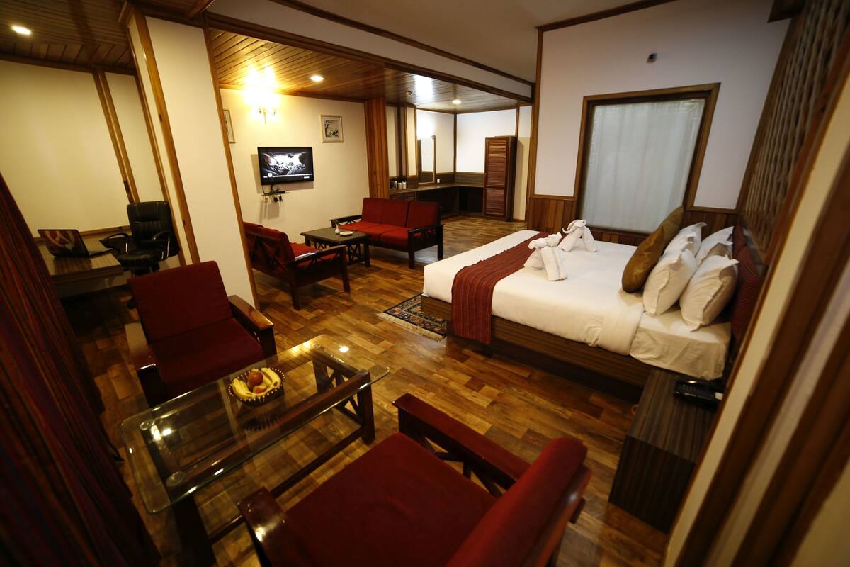 Quarto de hotel com uma cama, cadeiras vermelhas, uma mesa de centro de vidro, uma televisão e piso de madeira.