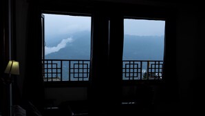 Quarto de hotel com cama, mesa de cabeceira e duas janelas com vista para montanhas e um lago.