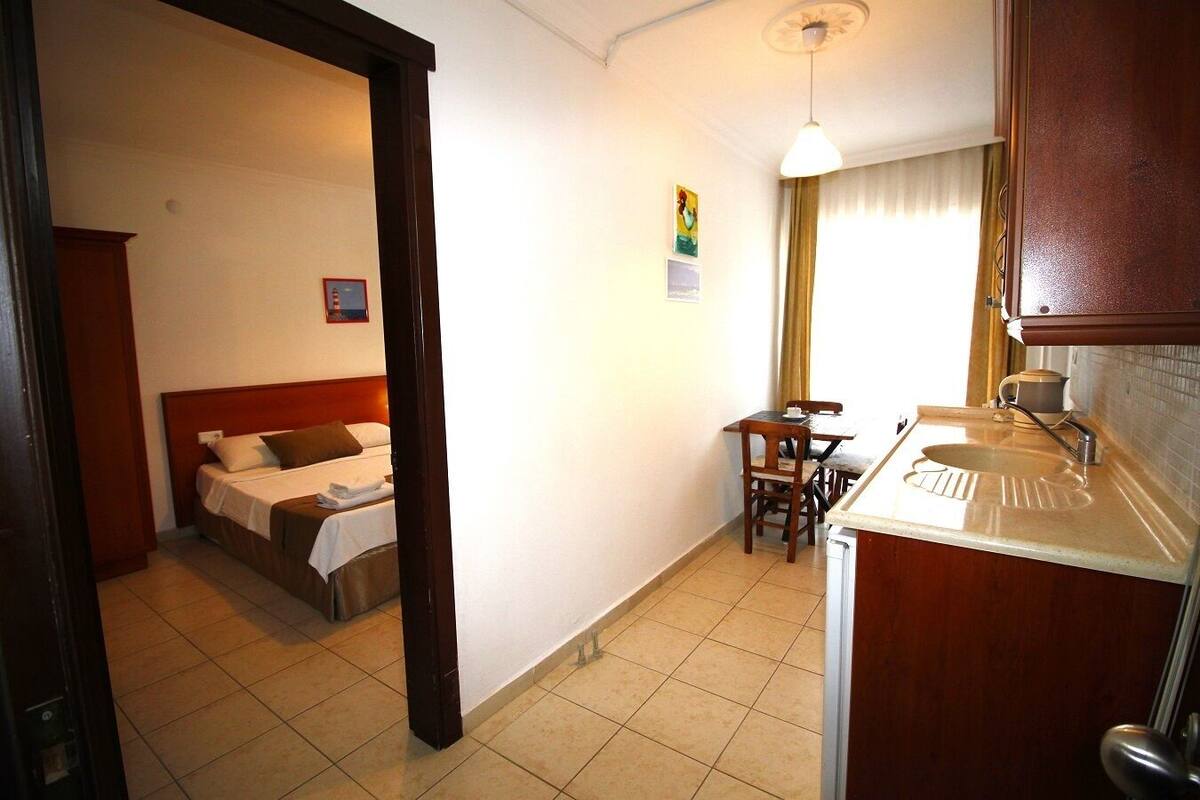 Quarto de hotel com cama, pequena mesa de jantar com cadeiras, cozinha equipada com pia e fogão, e janela com cortinas.