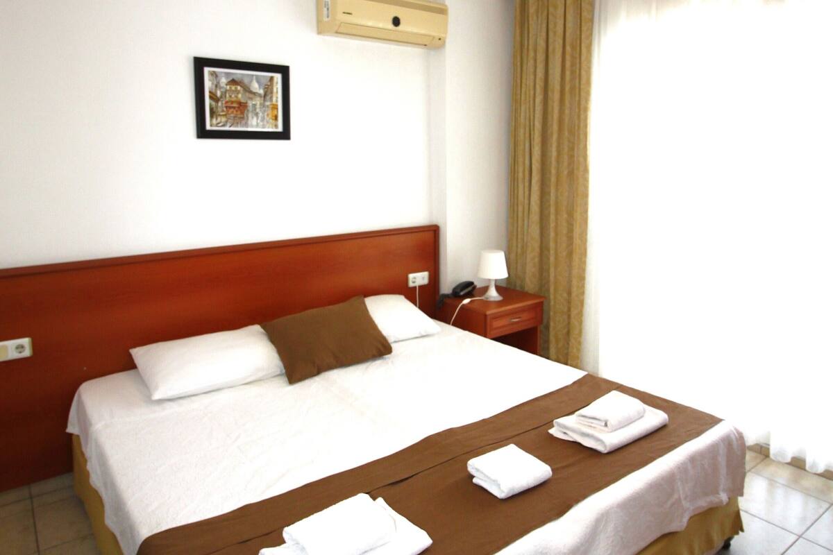 Quarto de hotel com uma cama grande, uma mesinha de cabeceira com abajur, um quadro na parede e uma janela com cortinas.