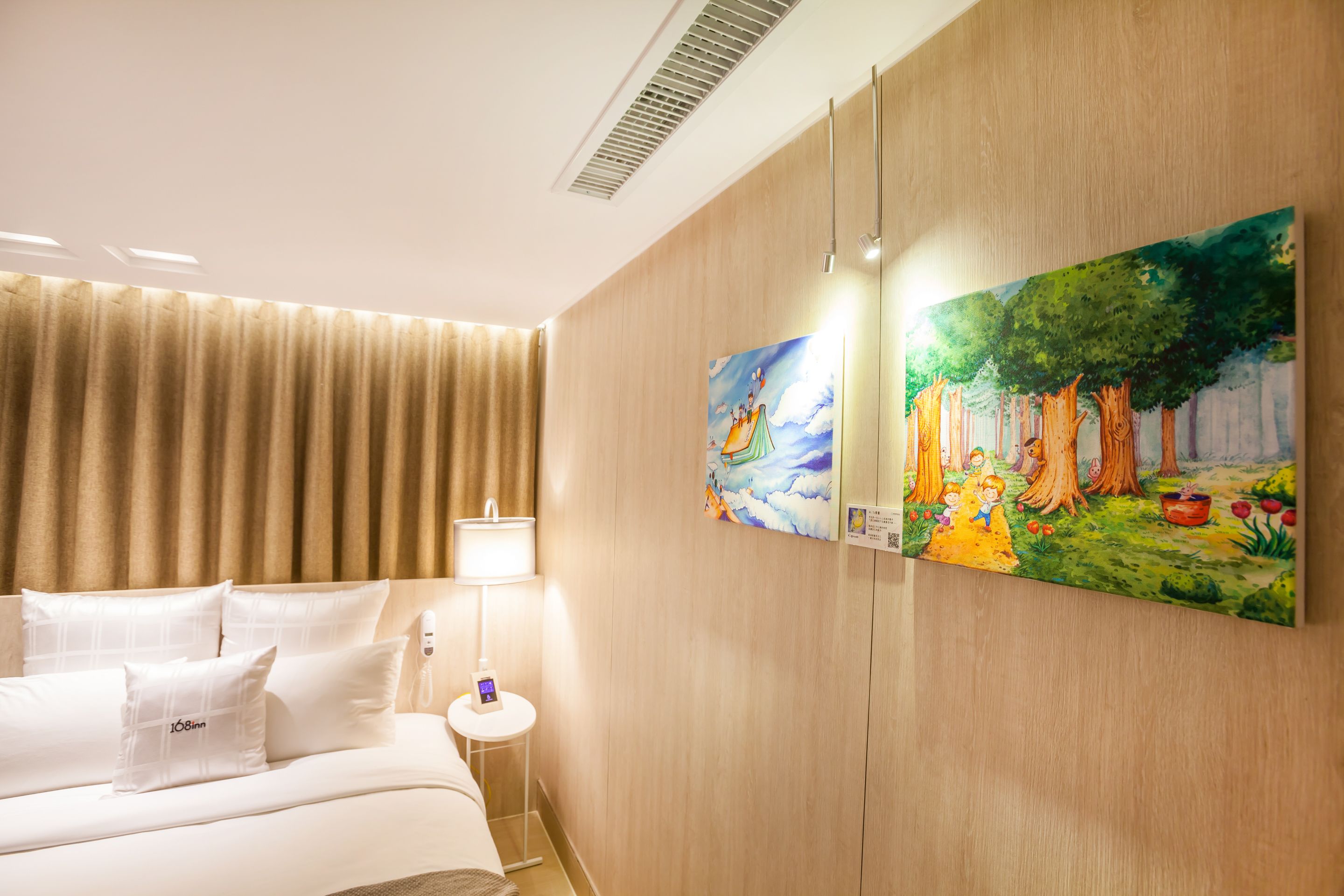 Quarto de hotel com uma cama, uma mesa de cabeceira, um abajur e duas pinturas na parede.