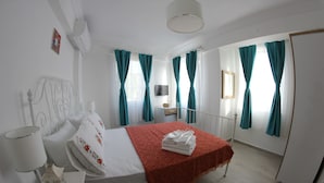 Quarto com cama, mesa de cabeceira com abajur, televisão na parede e cortinas cor turquesa.