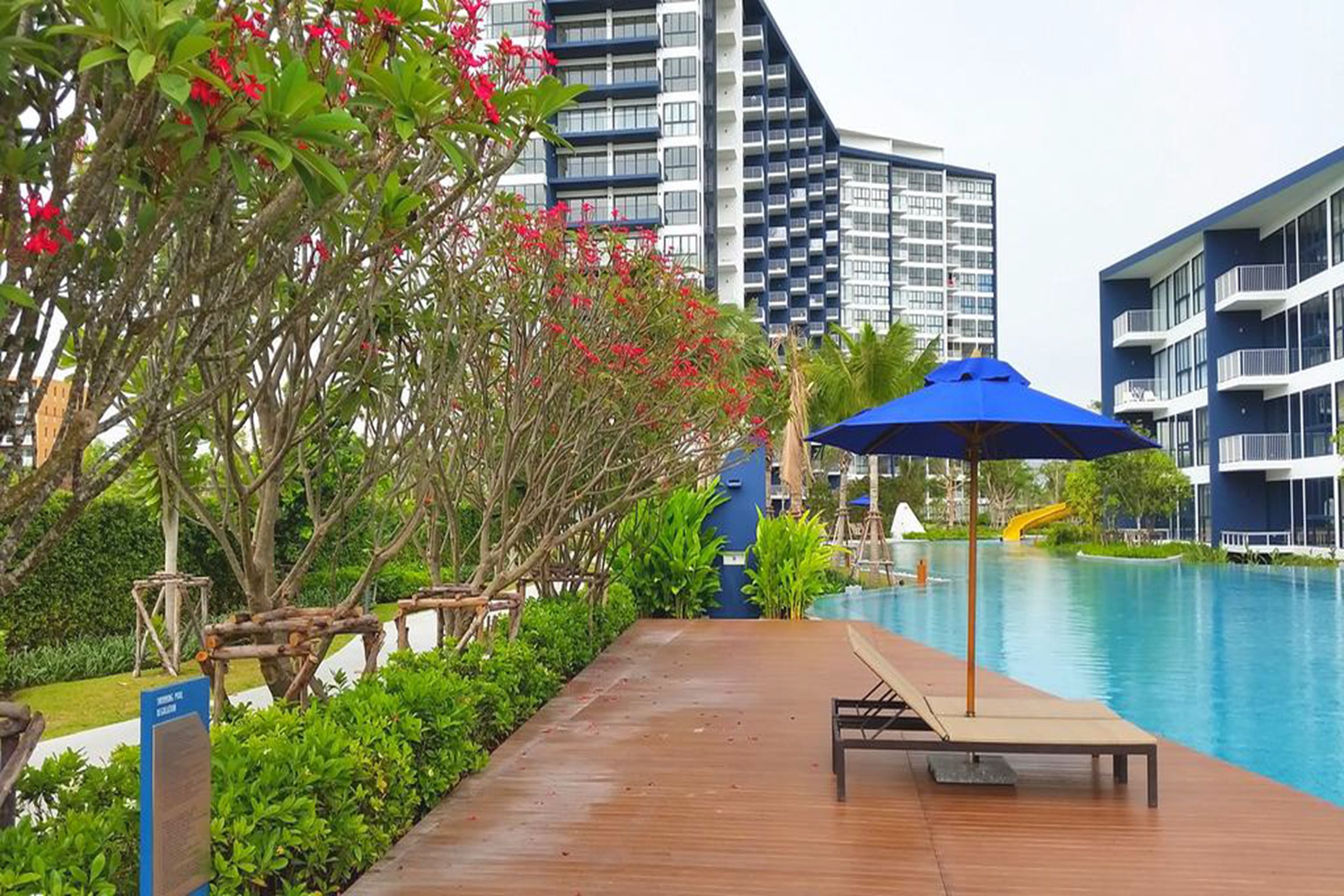 Um deck de madeira com espreguiçadeira e guarda-sol junto a uma piscina, cercado por vegetação e edifícios modernos.