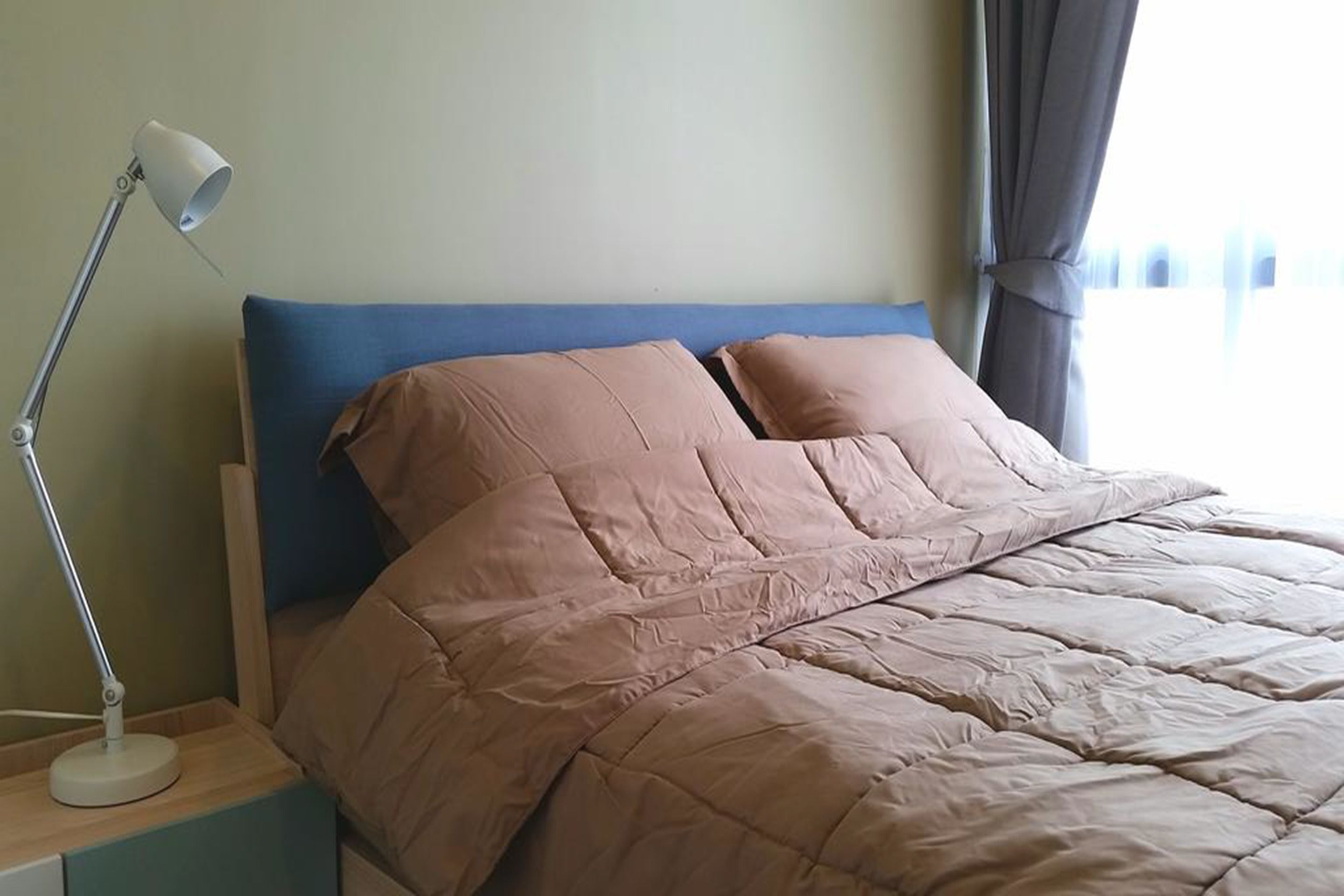 Uma cama bem arrumada com cabeceira azul, luminária ao lado e janela com cortinas.