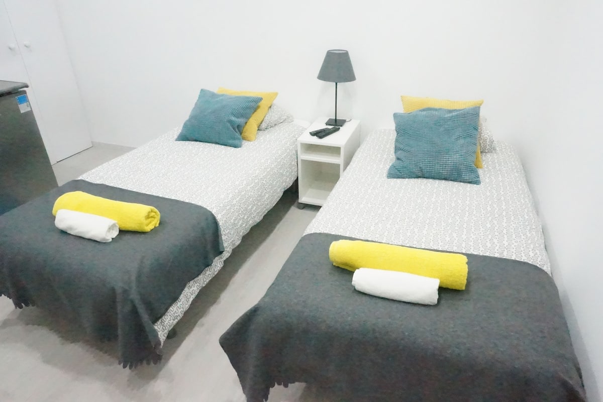 Quarto de hotel com duas camas individuais, cada uma com colcha estampada em branco e cinza, manta cinza escura e toalha amarela. Há uma mesa de cabeceira com abajur entre as camas.