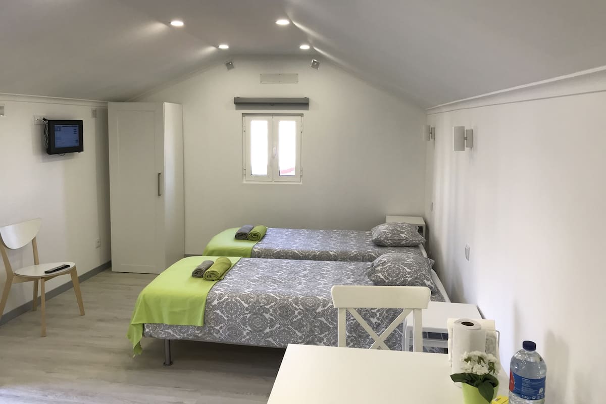 Quarto com duas camas, uma escrivaninha, uma cadeira, uma TV e uma janela.