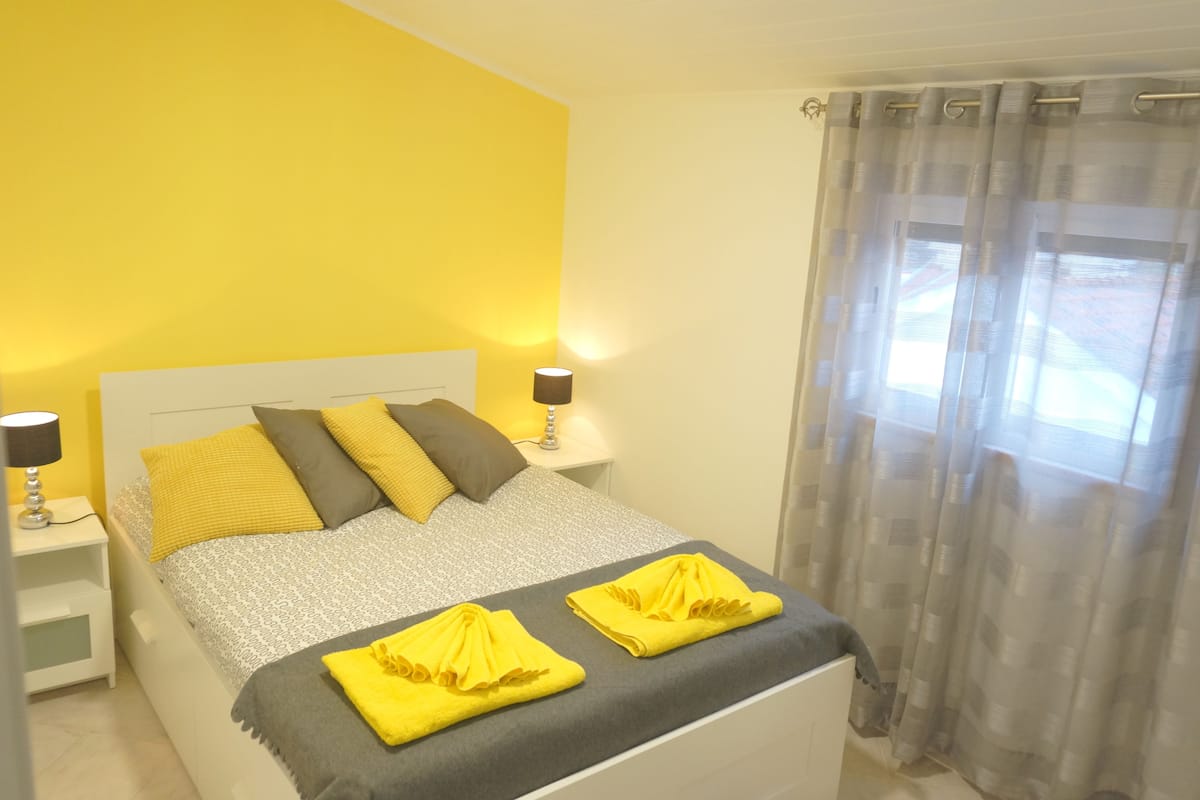 Uma cama bem arrumada com cabeceira cinza, duas luminárias de cabeceira e toalhas amarelas sobre a cama.